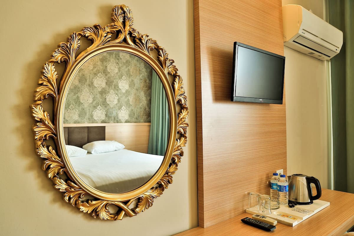 Quarto de hotel com um espelho grande e detalhado, cama com roupa de cama branca, televisão na parede e uma mesinha com chaleira, copos e controle remoto.