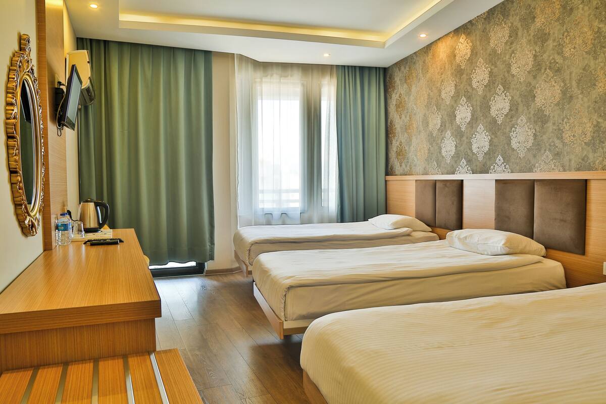 Quarto de hotel com duas camas de solteiro, uma escrivaninha de madeira, um espelho e uma janela com cortinas.