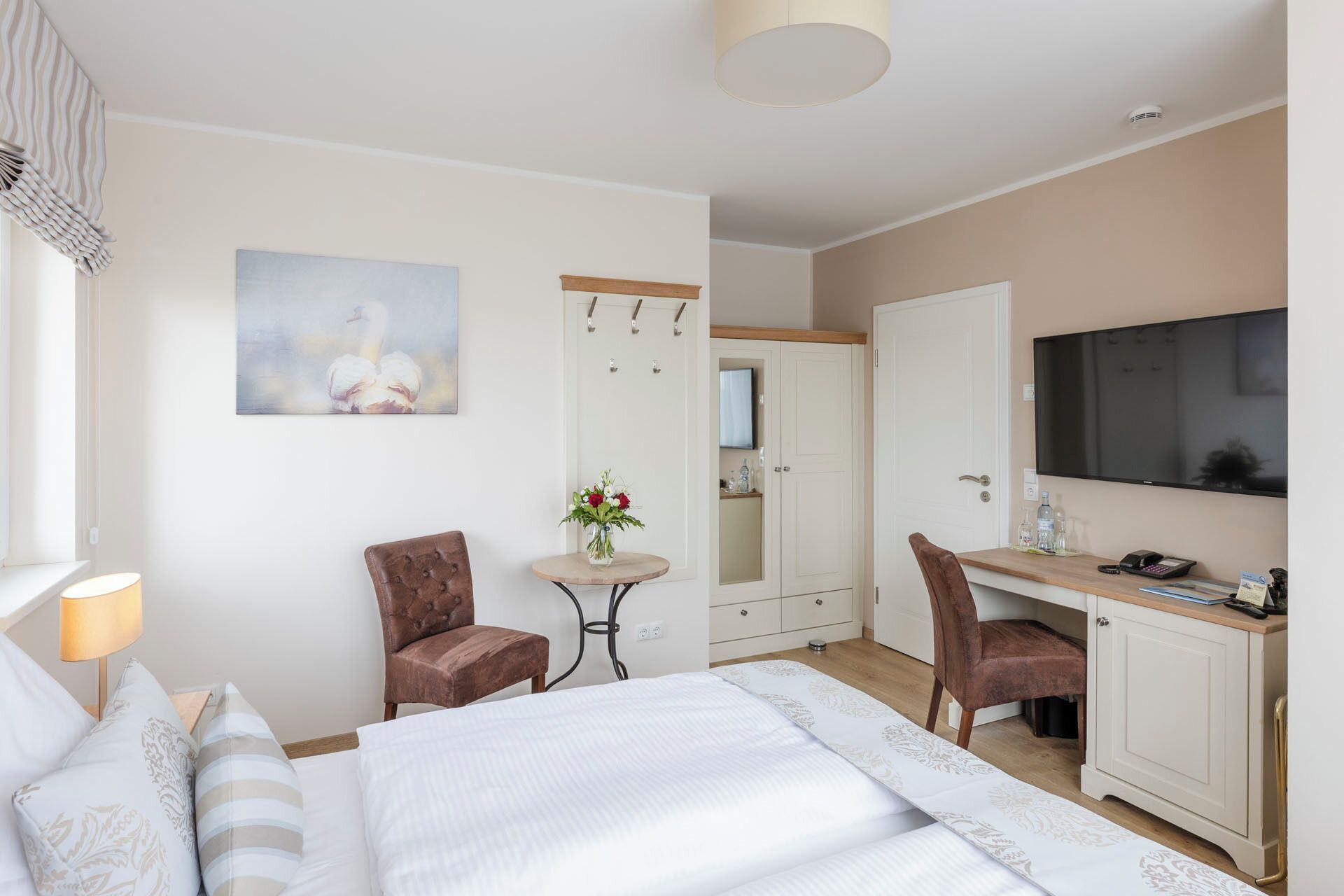 Quarto de hotel com uma cama, duas cadeiras, uma escrivaninha, uma televisão e um quadro na parede.