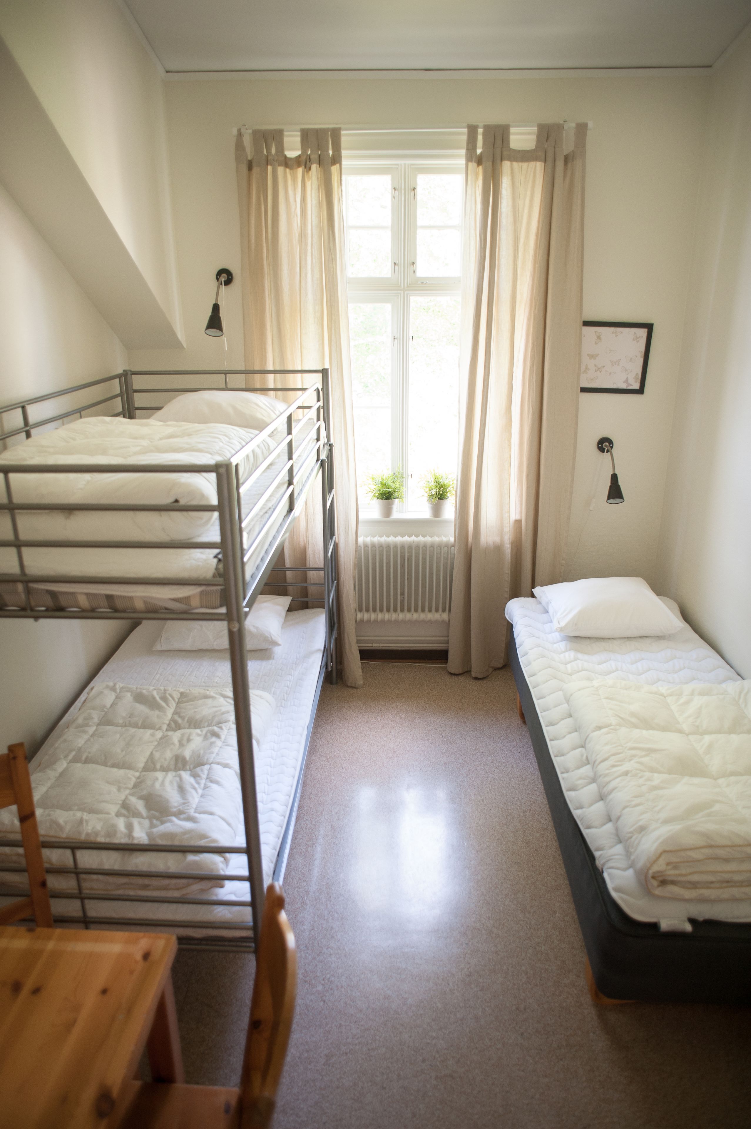 Quarto de dormitório com beliches, uma cama individual, janela com cortinas, radiador, escrivaninha e uma cadeira.