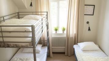 Quarto de dormitório com beliches, uma cama individual, janela com cortinas, radiador, escrivaninha e uma cadeira.