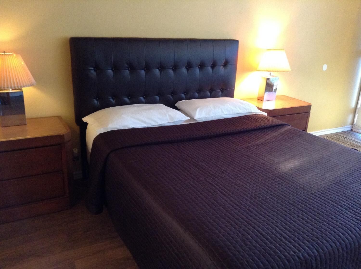 Uma cama bem arrumada com cabeceira escura, almofadas brancas e um cobertor escuro. Há duas mesas de cabeceira com abajures de cada lado da cama.