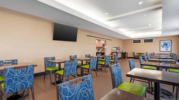 Uma área de refeições de hotel com mesas e cadeiras, uma televisão de tela plana na parede e uma cozinha ao fundo.