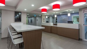 Uma área de recepção de hotel moderna, com um balcão branco, vários bancos altos e iluminação pendente vermelha.