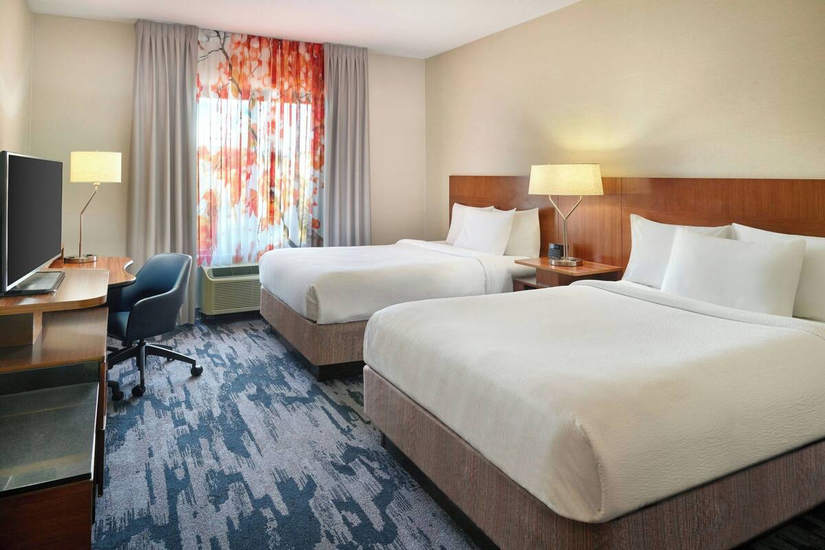 Quarto de hotel com duas camas, uma escrivaninha com computador, uma cadeira e uma janela grande com cortinas.