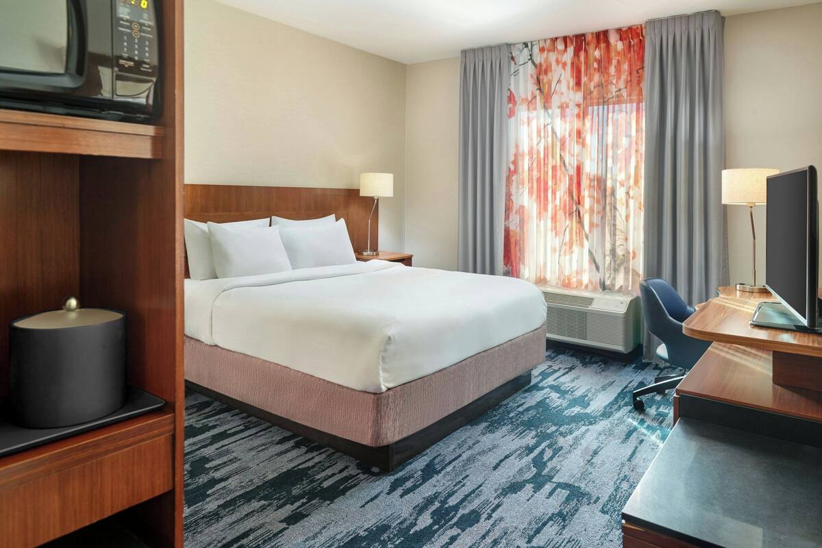 Quarto de hotel com cama, escrivaninha, cadeira, televisão, abajur e janela com cortinas.