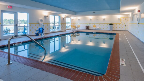 Piscina interior com água cristalina, cadeira de salva-vidas e área da piscina com cadeiras e toldo.