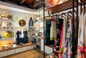 Quarto de hotel boutique com piso de madeira, divisória de vidro e diversos objetos, incluindo bolsas, chapéu e um cabideiro com roupas coloridas.
