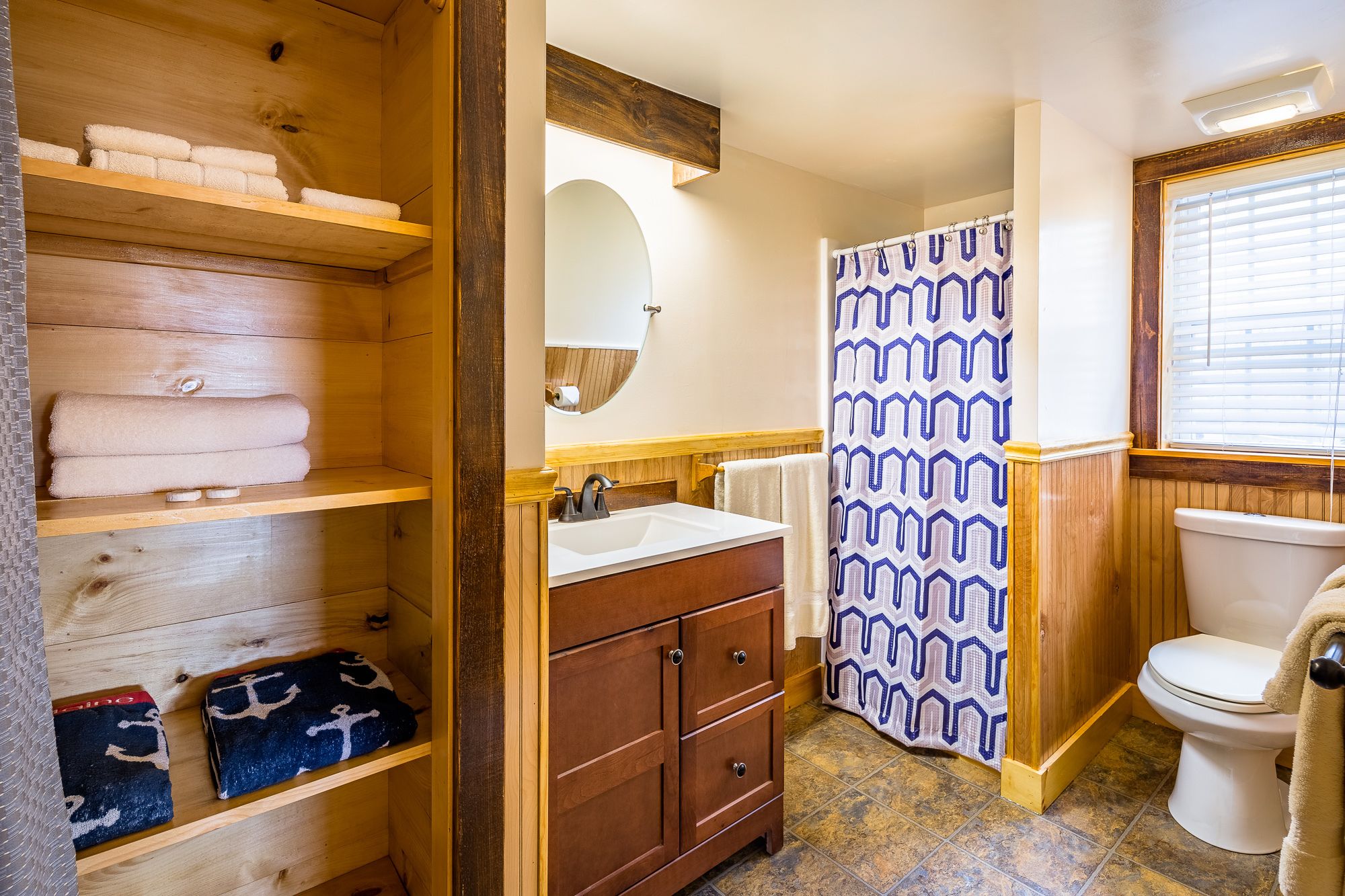 Casa de banho de cabana de madeira com uma prateleira de madeira com toalhas, uma pia com espelho, cortina de chuveiro estampada e sanita.