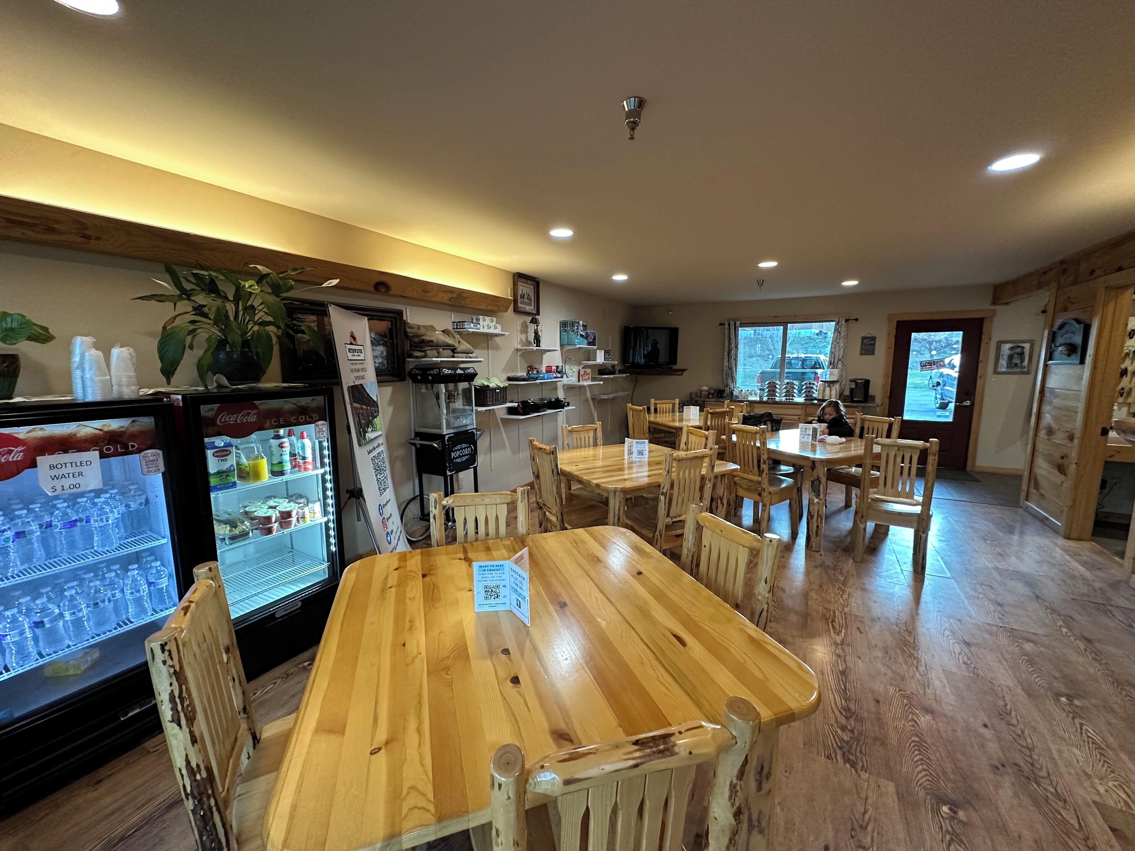 Um restaurante com mesas e cadeiras de madeira, um refrigerador de Coca-Cola, uma máquina de pipoca e uma janela com vista para o exterior.