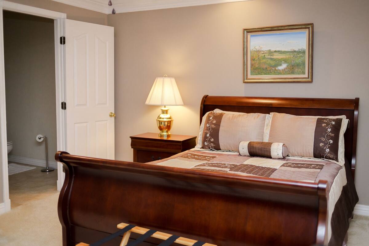 Um quarto com cama de madeira, mesa de cabeceira com abajur, pintura de paisagem emoldurada na parede e porta que dá para outro cômodo.