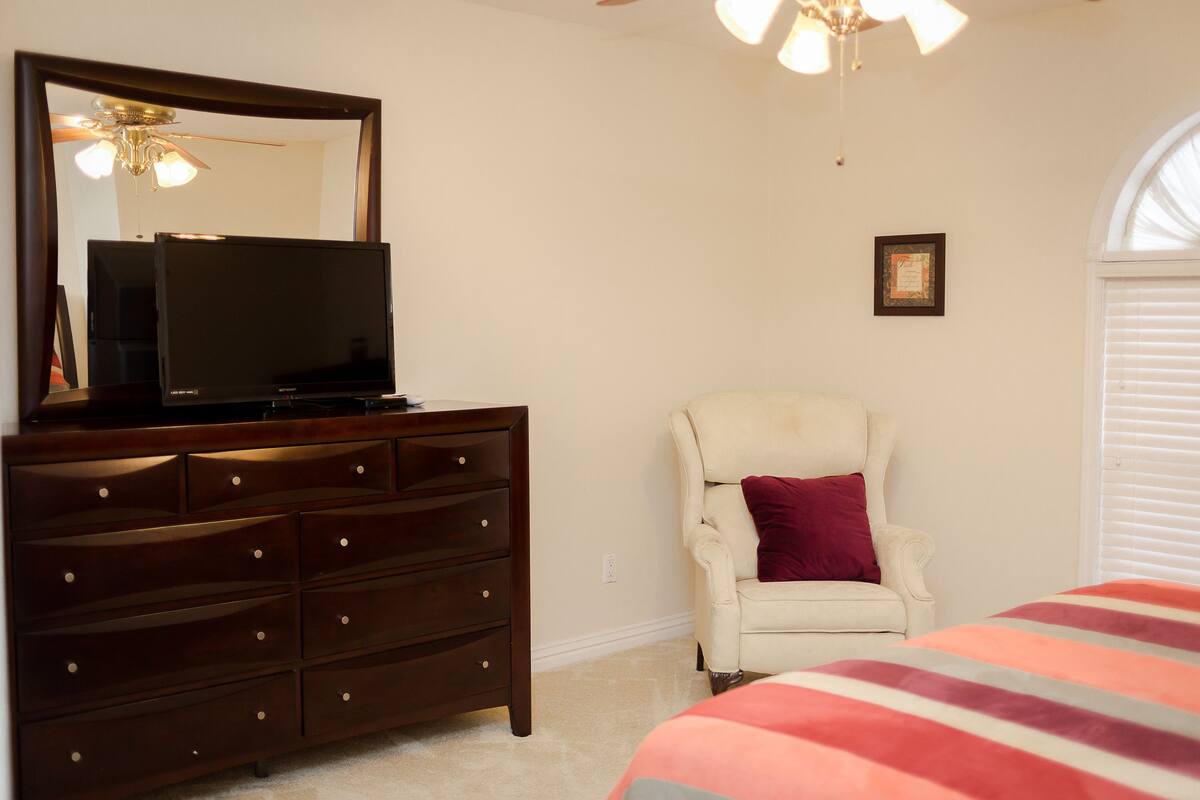 Um quarto com cômoda de madeira escura, poltrona branca, cama com colcha listrada, ventilador de teto e um quadro na parede.