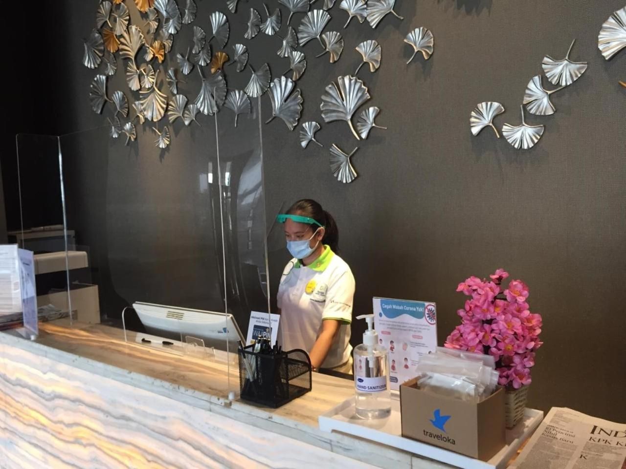 Bancada de recepção com dispensador de álcool em gel, placa com avisos sobre precauções para COVID-19 e parede decorativa com padrões semelhantes a folhas.