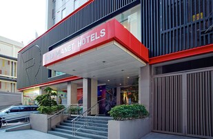 Entrada de hotel moderna com uma placa chamativa, escadaria e acesso por portão.