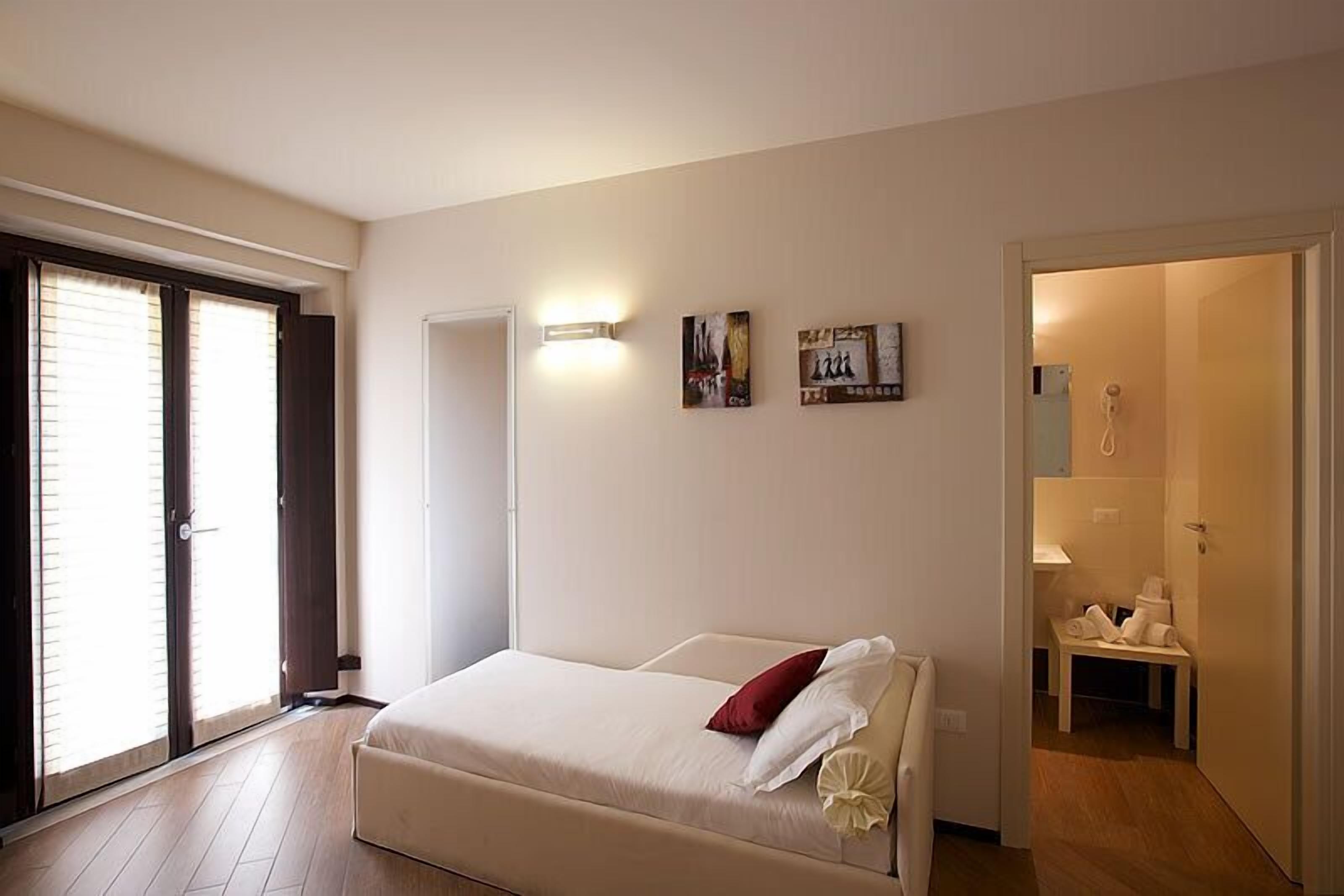 Quarto com cama, porta, luminária na parede e uma mesinha com toalhas.