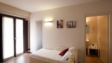 Quarto com cama, porta, luminária na parede e uma mesinha com toalhas.