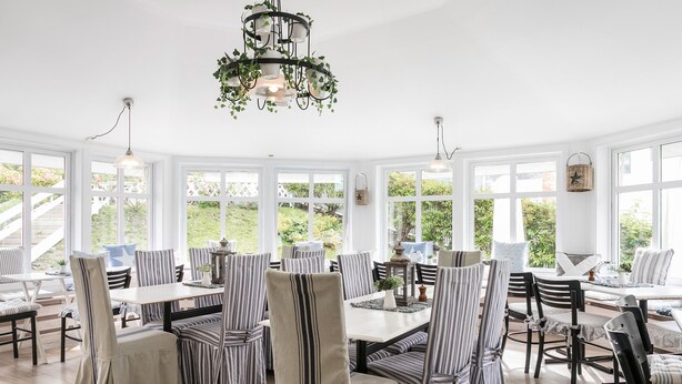 Uma sala de jantar luminosa com cadeiras estofadas em listras e mesas, janelas amplas e um lustre.