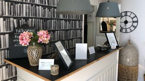 Bancada de recepção com uma lousa exibindo uma mensagem de boas-vindas, uma placa emoldurada e um pequeno vaso com flores cor-de-rosa. Atrás do balcão, há uma estante cheia de livros.