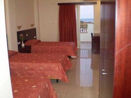 Quarto de hotel com três camas, cabeceira de madeira, mesa de cabeceira com um vaso e vista para o mar através de uma porta de varanda.