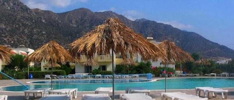 Área de piscina com espreguiçadeiras e guarda-sóis de palha, cercada por muros de pedra e montanhas ao fundo.