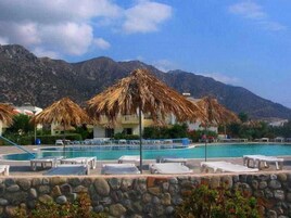 Área de piscina com espreguiçadeiras e guarda-sóis de palha, cercada por muros de pedra e montanhas ao fundo.