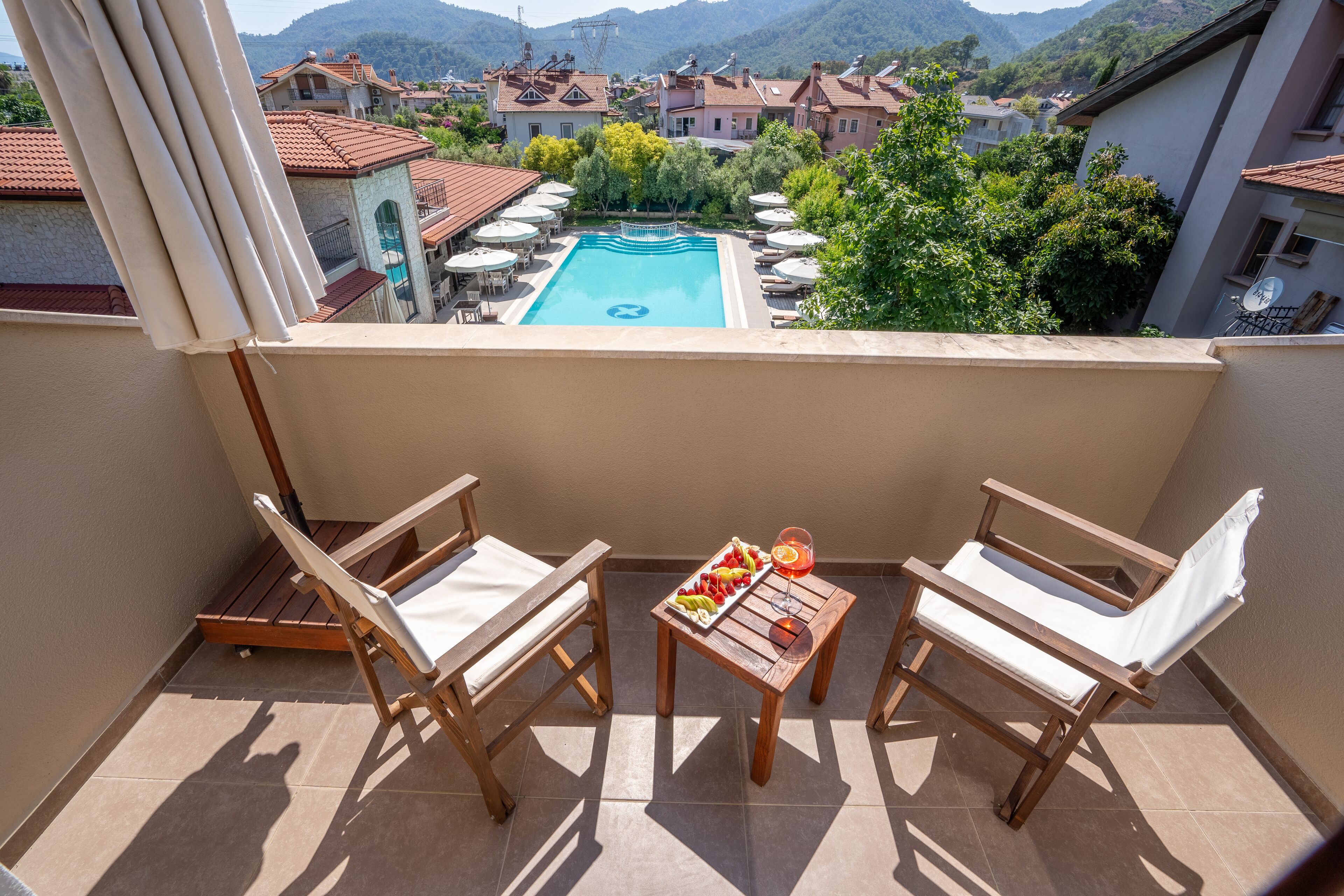 Uma varanda com duas cadeiras de madeira, uma mesa pequena com uma travessa de frutas e um copo de bebida, e vista para uma piscina e edifícios ao redor.