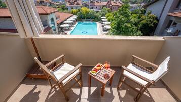 Uma varanda com duas cadeiras de madeira, uma mesa pequena com uma travessa de frutas e um copo de bebida, e vista para uma piscina e edifícios ao redor.