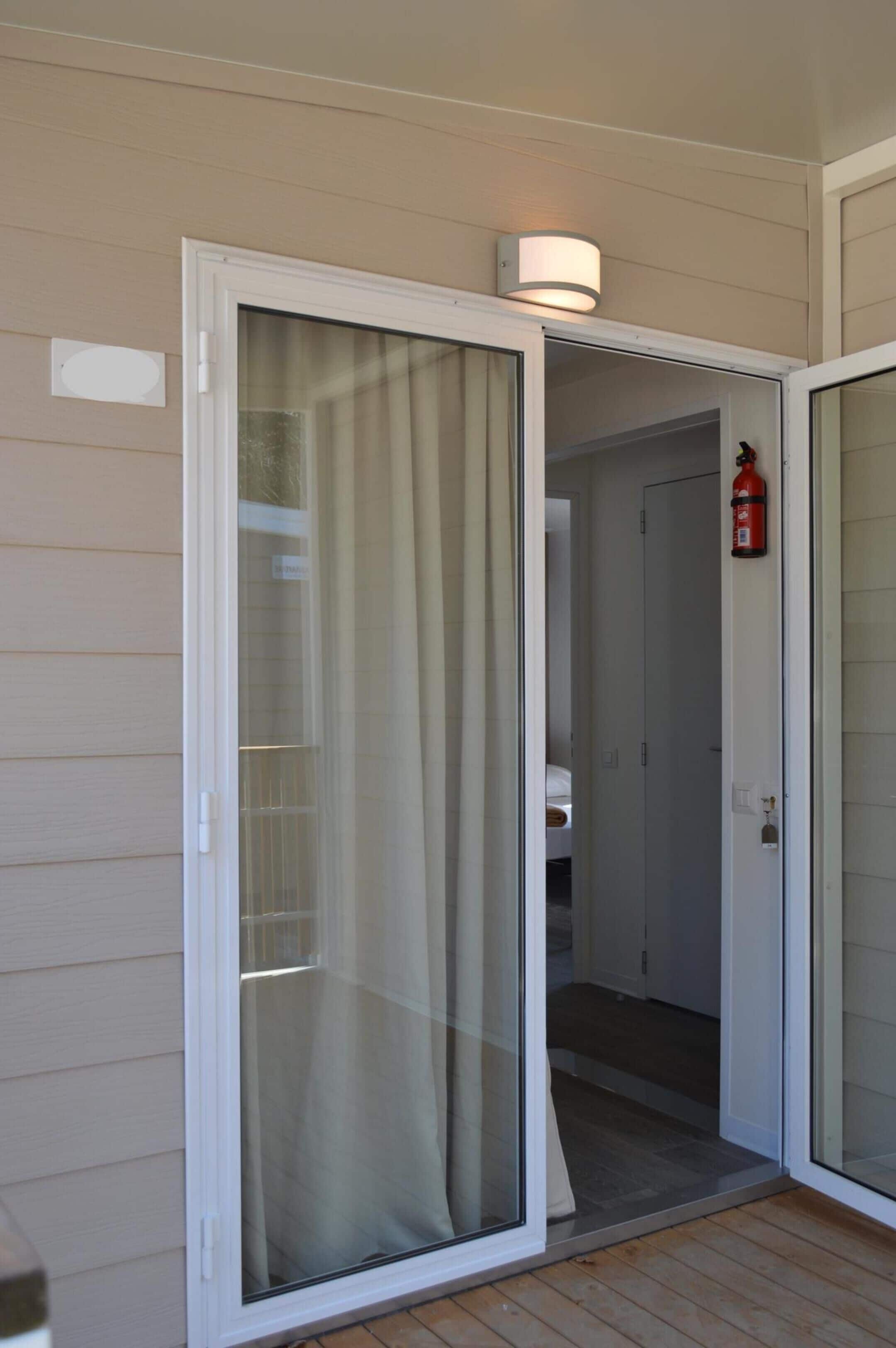 Quarto com porta de correr de vidro, extintor fixado na parede e uma cama visível ao fundo.
