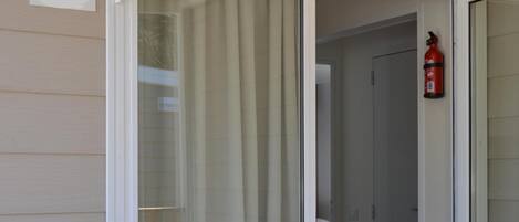 Quarto com porta de correr de vidro, extintor fixado na parede e uma cama visível ao fundo.