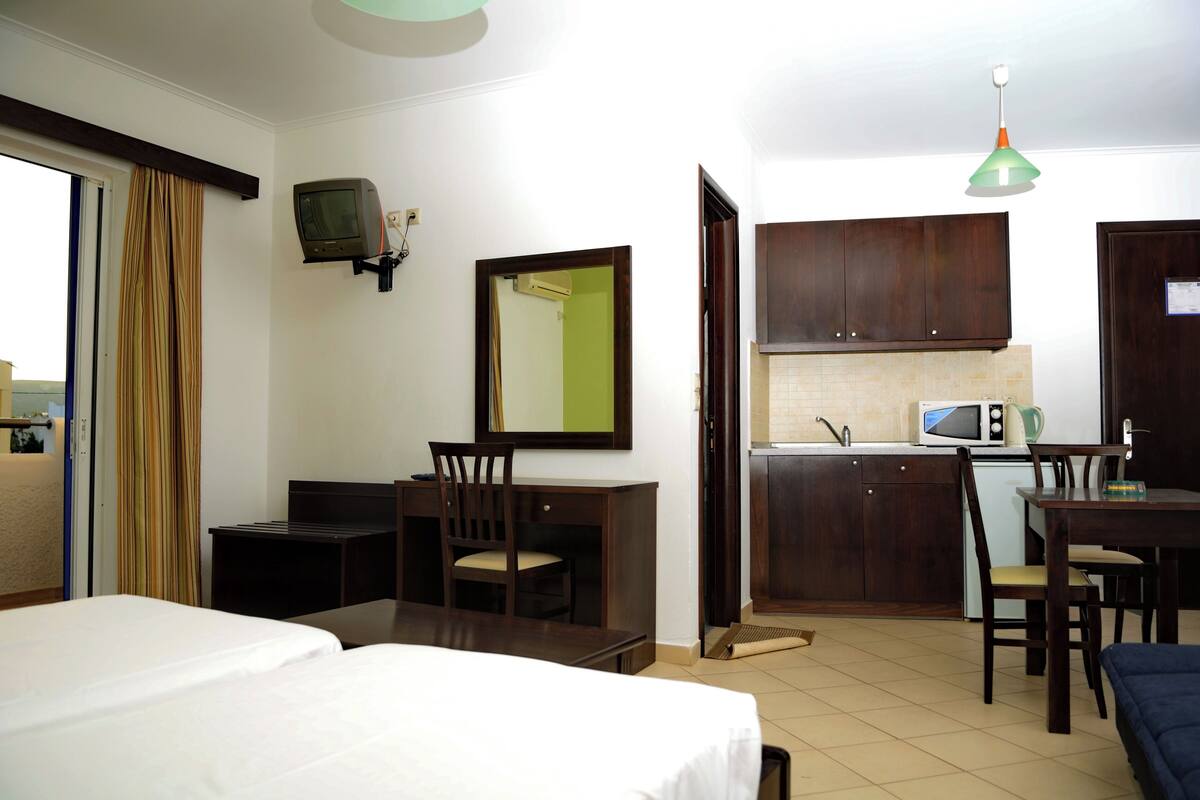 Quarto de hotel compacto com cama, escrivaninha com cadeira, cozinha compacta e banheiro.