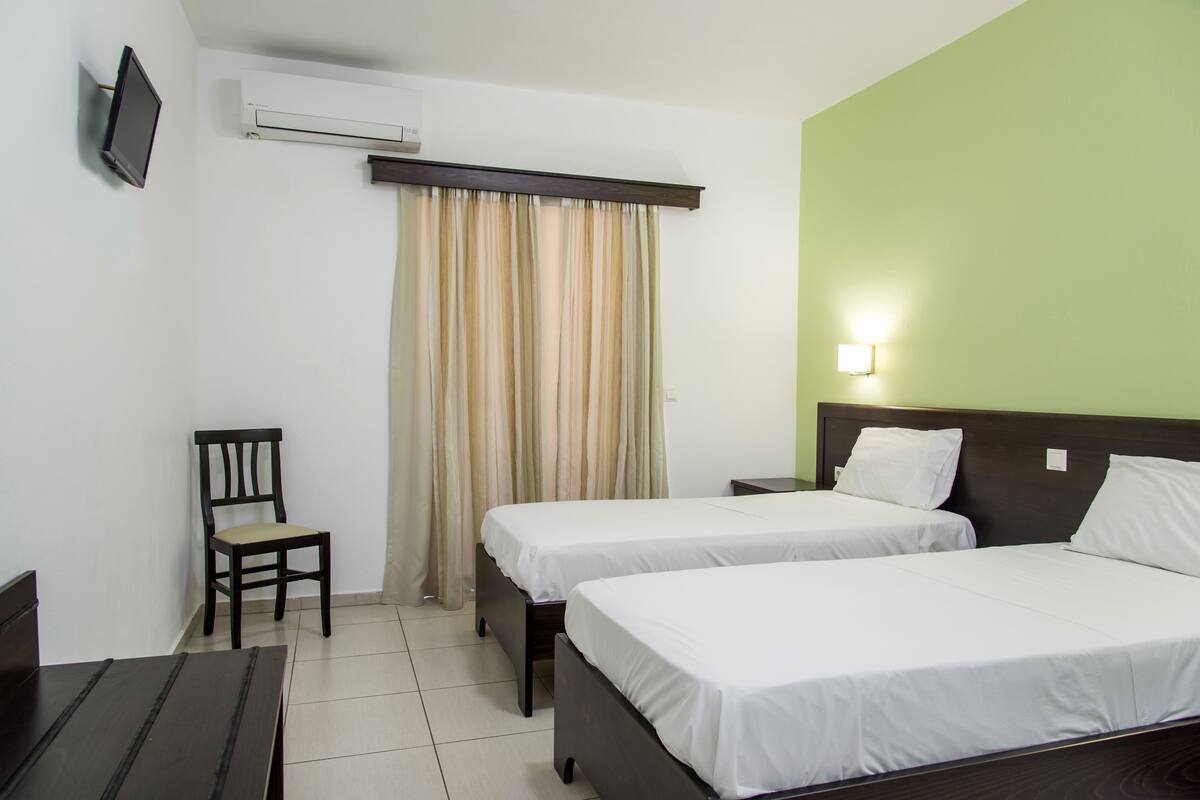 Quarto de hotel com duas camas de solteiro, uma escrivaninha, uma cadeira, uma televisão e um ar condicionado.