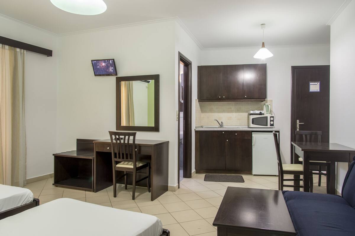 Quarto de hotel compacto com cama, escrivaninha com cadeira, cozinha compacta equipada com micro-ondas e televisão.