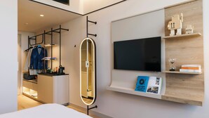 Quarto de hotel moderno com uma cama, televisão, espelho, guarda-roupa e uma estante com um livro e objetos decorativos.