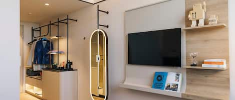Quarto de hotel moderno com uma cama, televisão, espelho, guarda-roupa e uma estante com um livro e objetos decorativos.