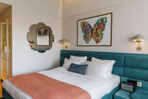 Cama bem arrumada com roupa de cama branca e laranja, cabeceira azul-turquesa e luminária fixada na parede.