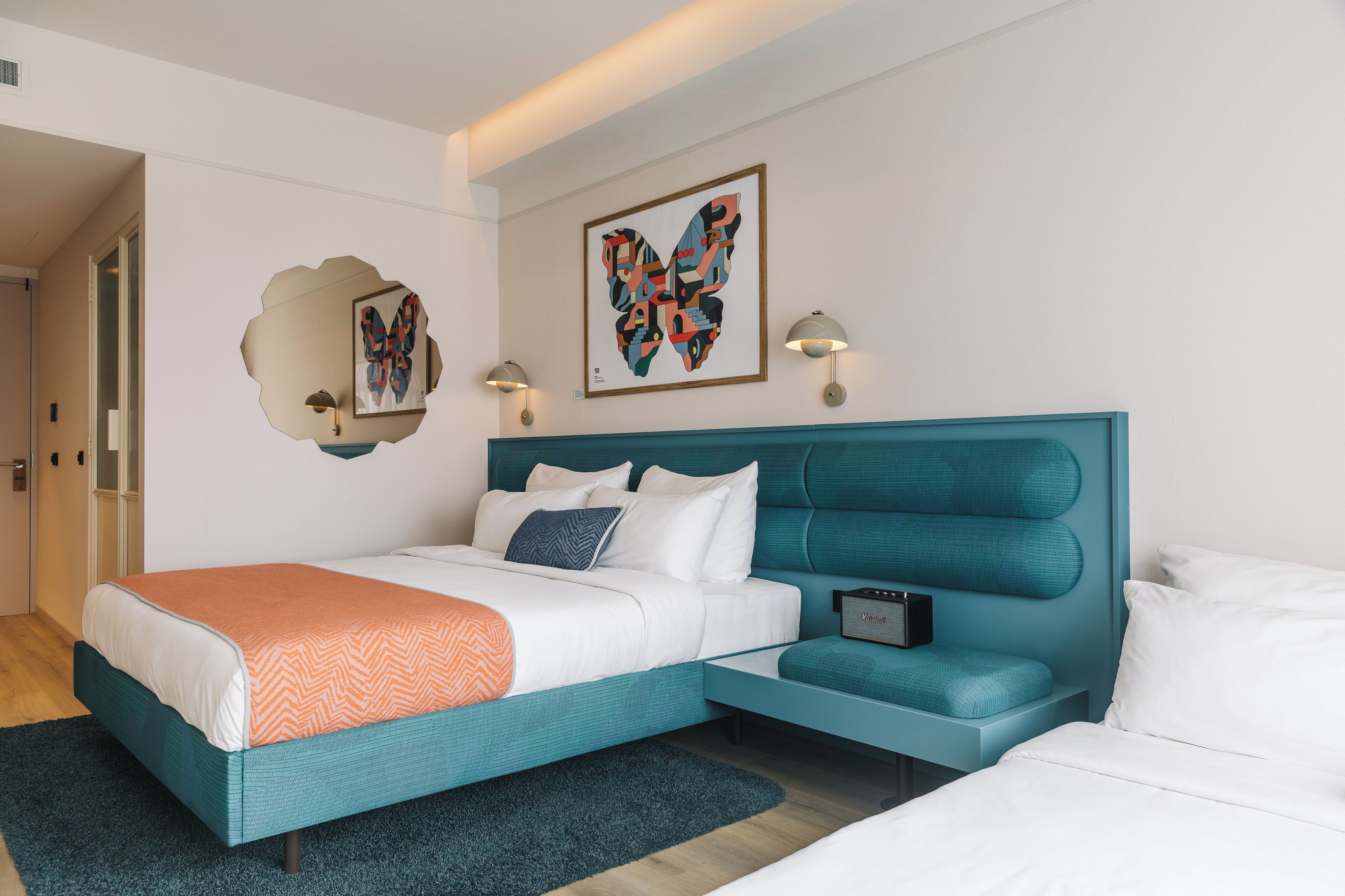 Quarto de hotel moderno com uma parede de destaque azul-turquesa, uma cama grande com roupa de cama branca, uma mesa lateral e duas luminárias fixadas na parede.