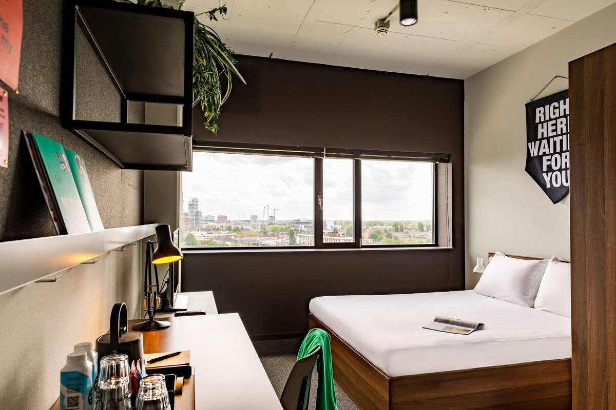 Quarto de hotel moderno com cama, escrivaninha e uma grande janela com vista para a cidade.