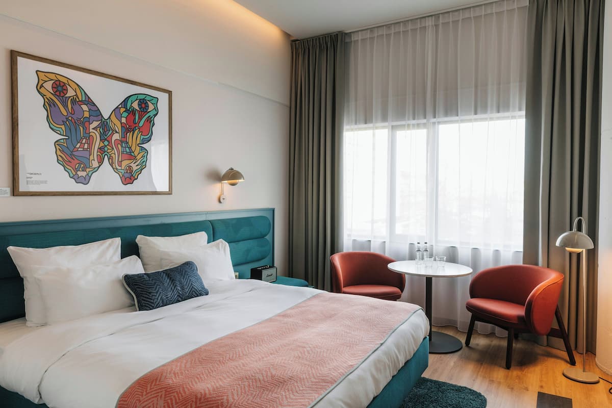 Quarto de hotel com cama, duas cadeiras, uma mesinha, um abajur, janela com cortinas e uma pintura de borboleta na parede.