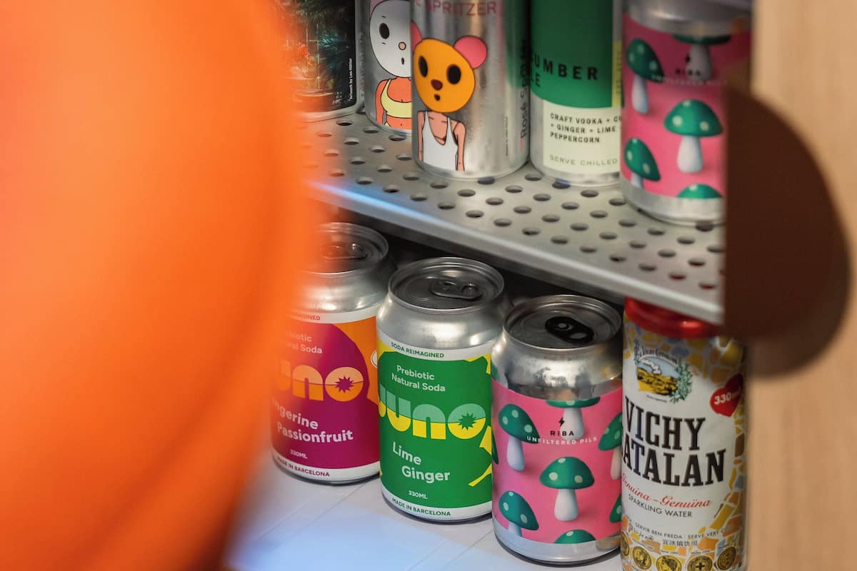 Uma estante com várias latas de bebidas, incluindo marcas como Prizer, Dujon e Vichy Catalan.