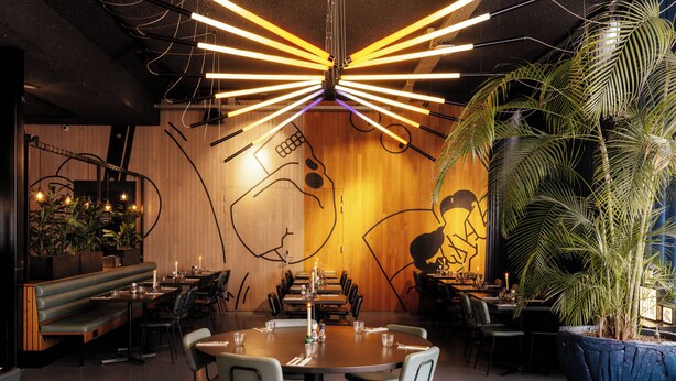 Um restaurante com uma mesa redonda central, diversas opções de assento e uma grande planta em vaso.