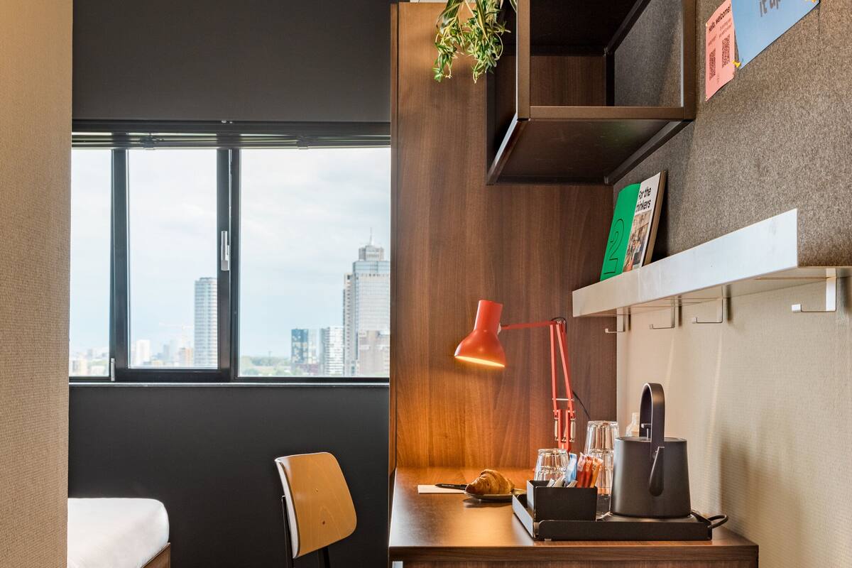 Quarto de hotel moderno com cama, escrivaninha, cadeira, planta em vaso e uma vista da cidade através de uma janela grande.