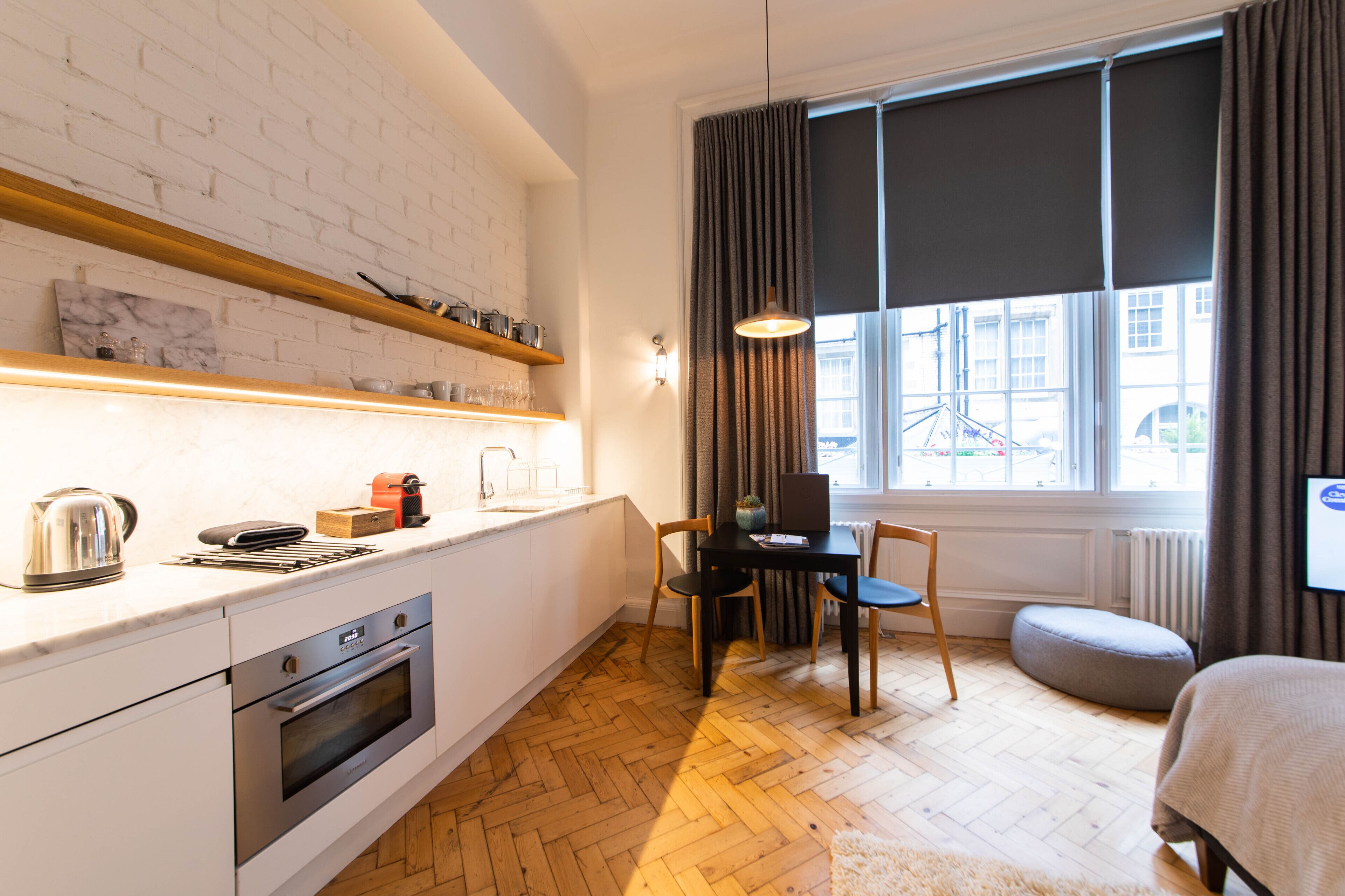 Uma cozinha moderna com armários brancos, forno embutido e bancada de madeira. Há uma área de refeições com mesa e cadeiras, e uma janela grande com cortinas.