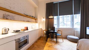 Uma cozinha moderna com armários brancos, forno embutido e bancada de madeira. Há uma área de refeições com mesa e cadeiras, e uma janela grande com cortinas.