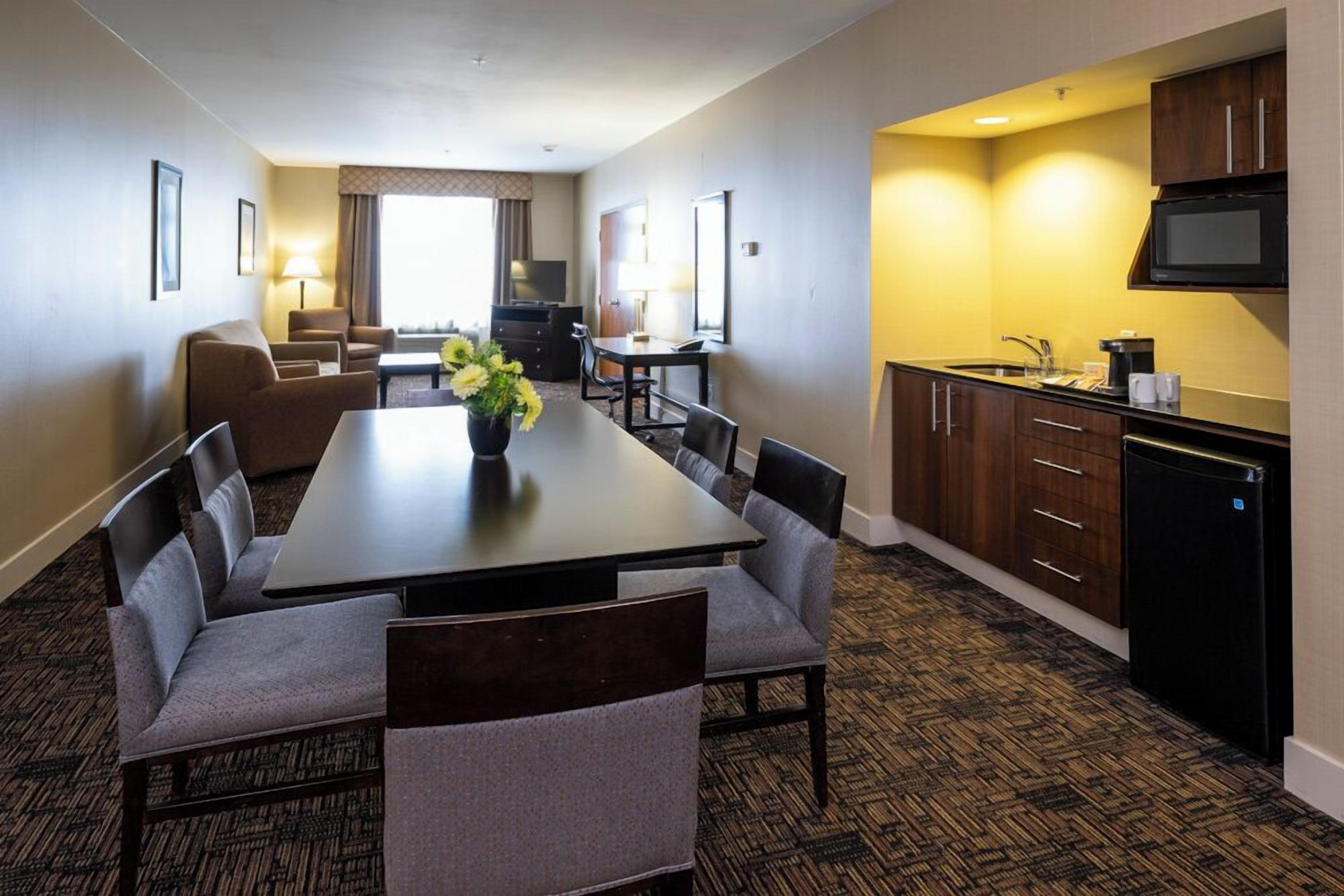 Quarto de hotel com mesa de jantar, cadeiras, televisão, cozinha compacta e piso coberto de carpete.