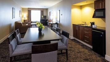 Quarto de hotel com mesa de jantar, cadeiras, televisão, cozinha compacta e piso coberto de carpete.