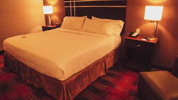 Quarto de hotel com uma cama grande, duas mesas de cabeceira, um criado-mudo, uma cadeira e piso coberto por carpete.