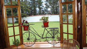 Janela de madeira com bicicleta e plantas em vasos no interior, com vista para um jardim gramado.