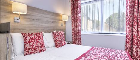 Quarto de hotel com uma cama grande, duas luminárias fixadas na parede, uma janela com cortinas e uma cabeceira de madeira.