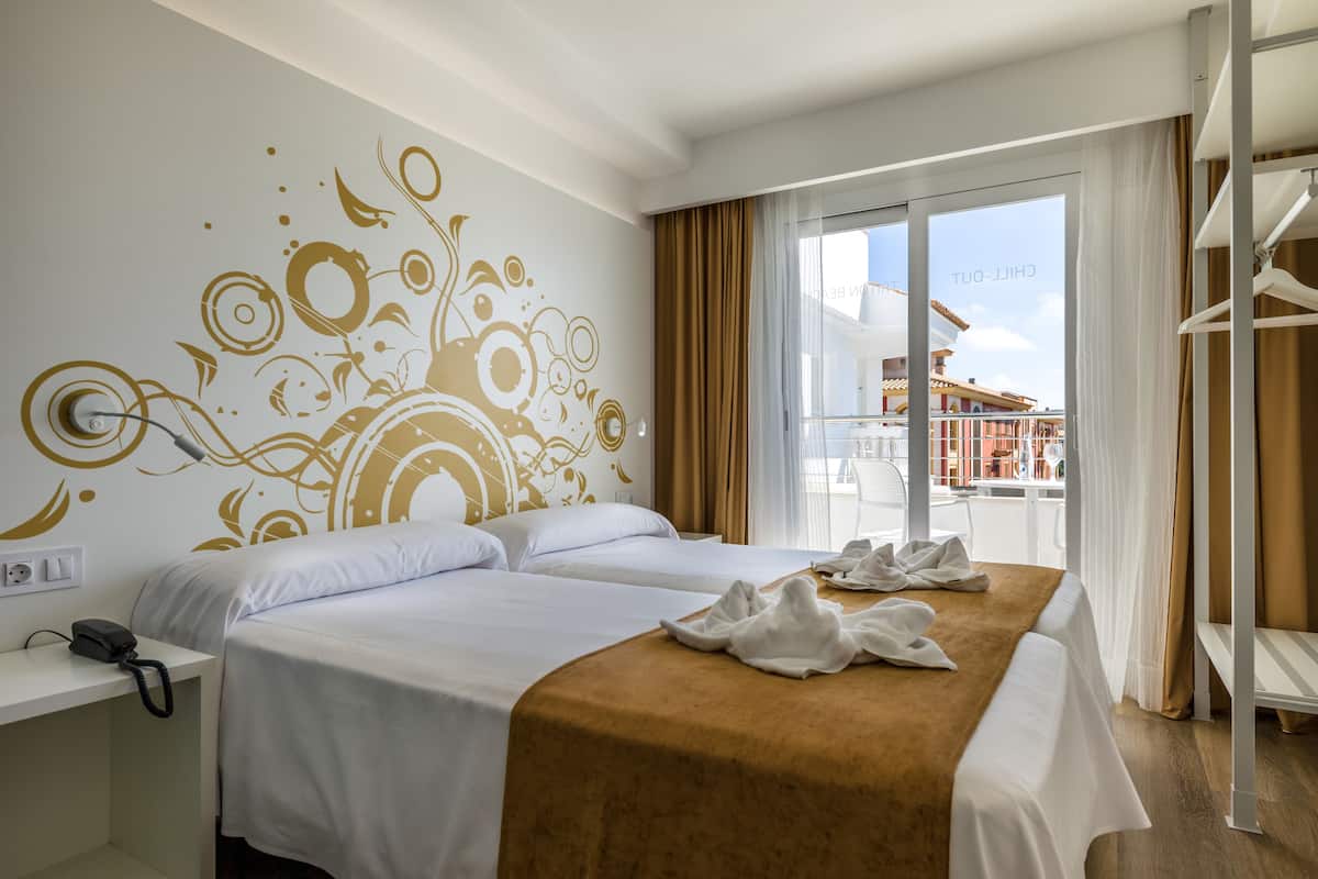 Quarto de hotel com duas camas, uma janela grande com cortinas, uma parede com padrões decorativos e vista para edifícios.