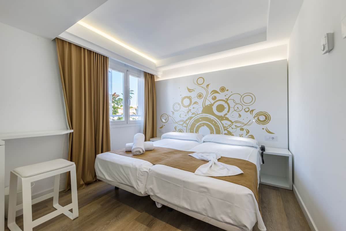 Quarto de hotel com cama, uma mesinha branca, criado-mudo e um detalhe decorativo na parede.